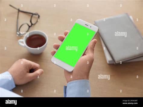 Man holding smartphone with green screen at wooden table indoors, closeup. Gadget display with ...